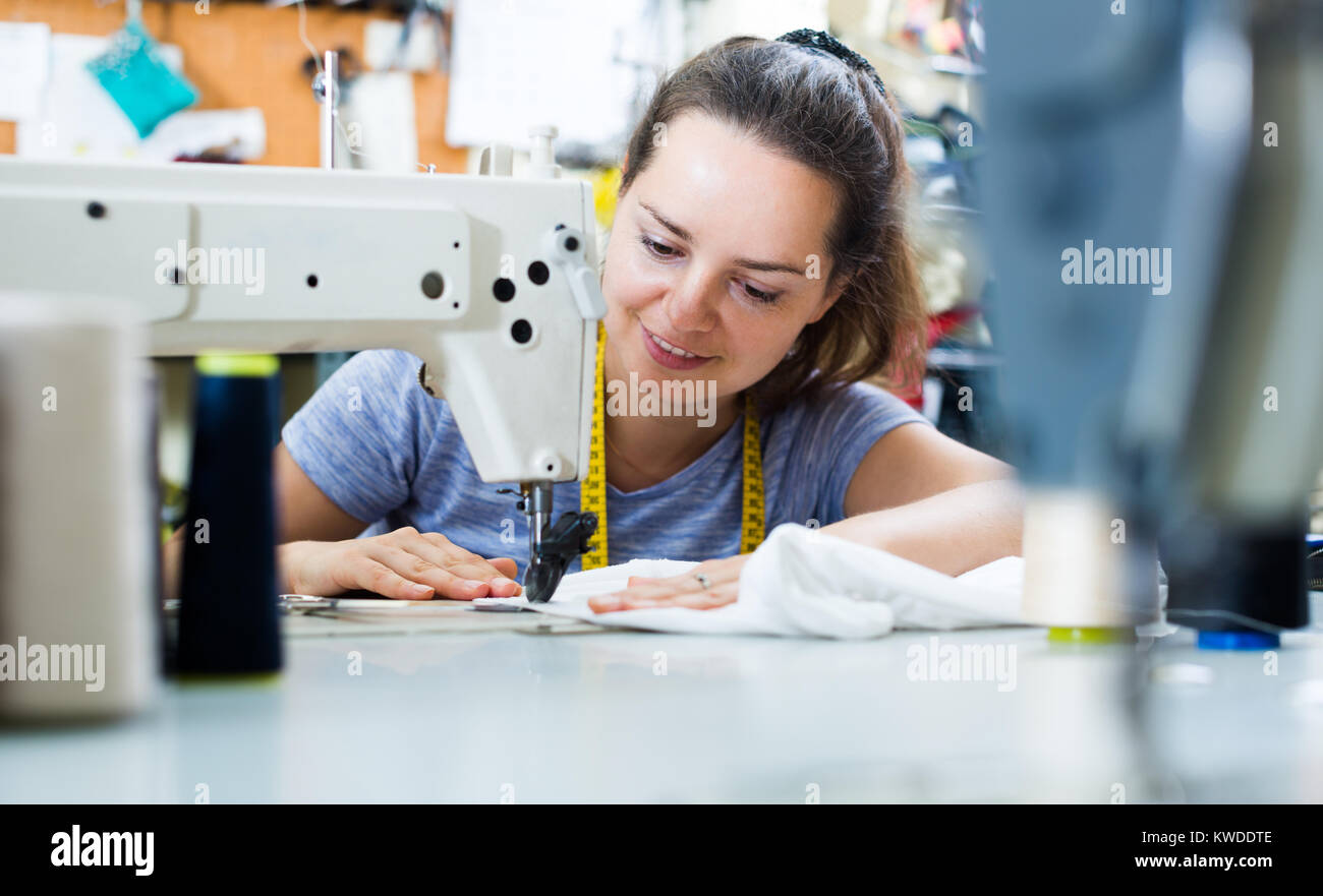 Positive young woman sewing with professional machine at workshop Stock ...