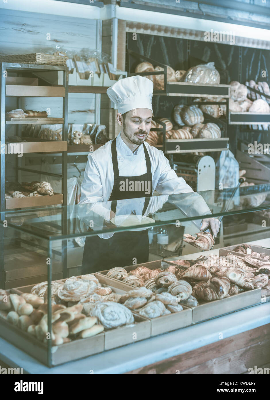 Cheerful positive man baker showing warm tasty croissant in bakery ...