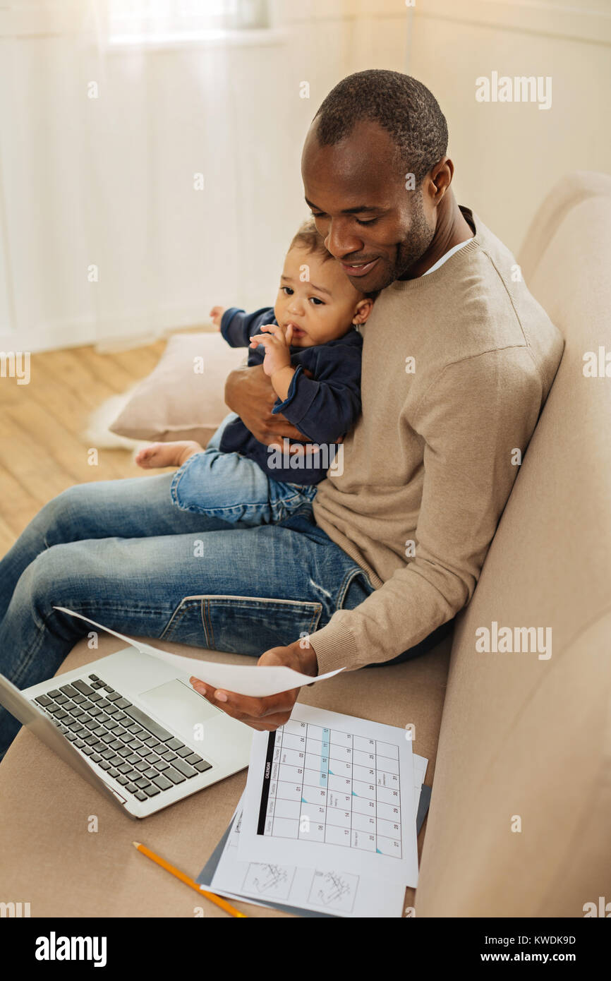 Smiling father working on the laptop and holding his son Stock Photo ...