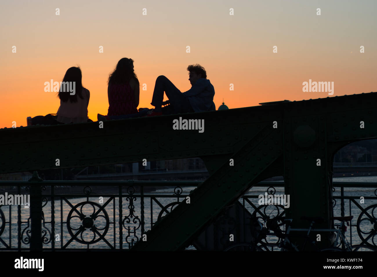 Silhouettes of tree young people sitting and talking on bridge beam ...