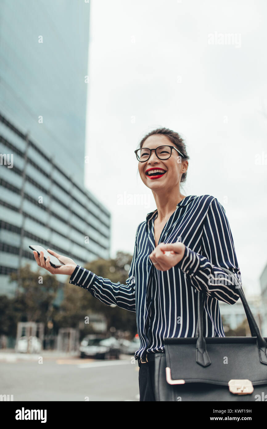 Smiling female business professional holding cell phone and hailing ...