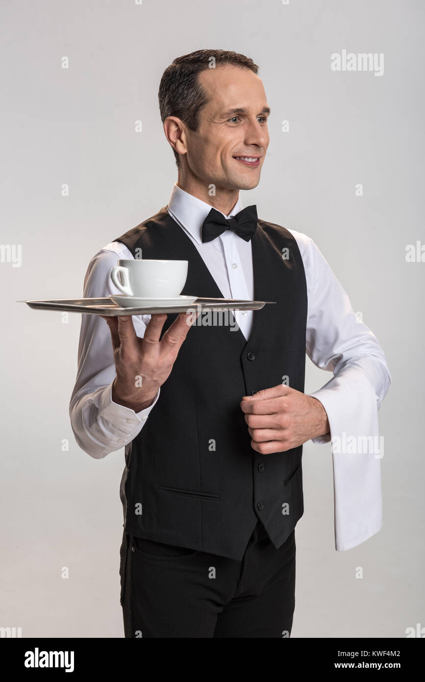 Your coffee. Jolly pensive male waiter posing on the grey background ...