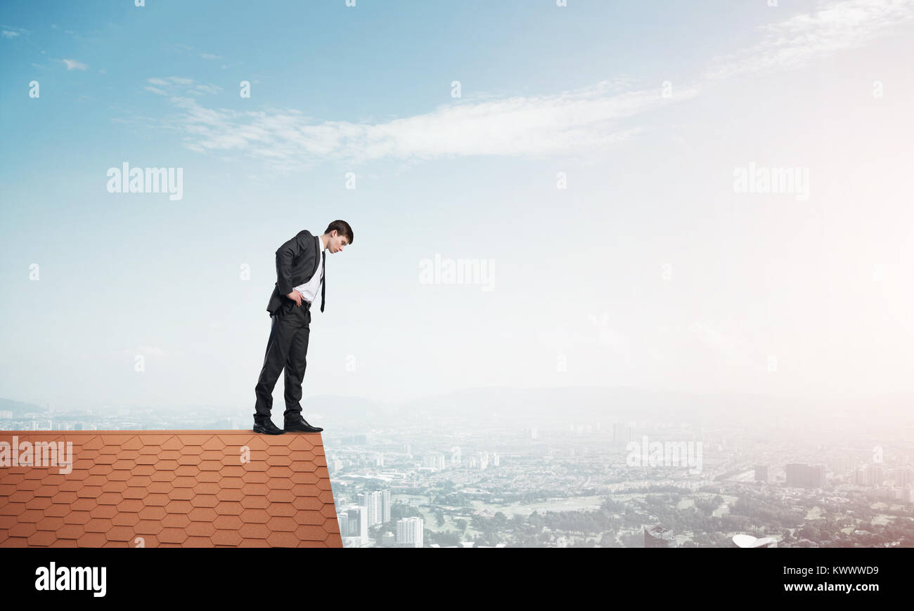 Young businessman standing on edge of house brick roof and looking down ...