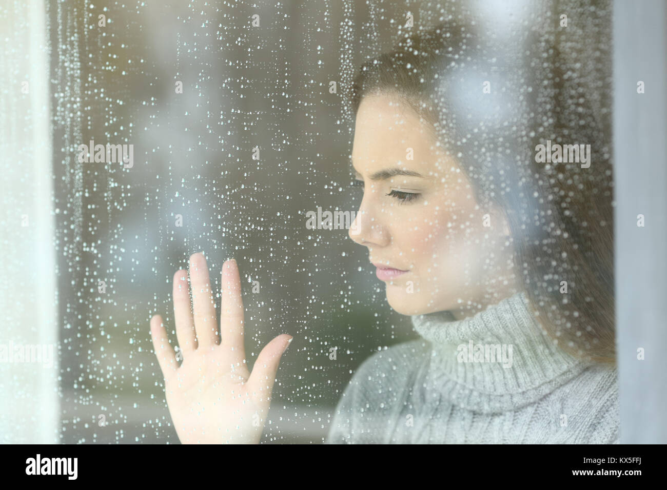 Portrait of a sad girl looking through a window in a rainy day at home ...