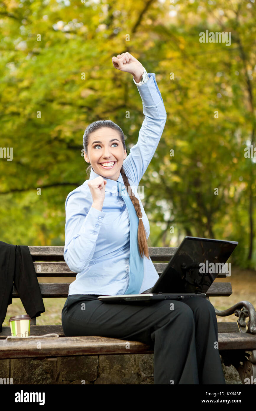 Excited businesswoman sitting on a park bench with laptop computer ...