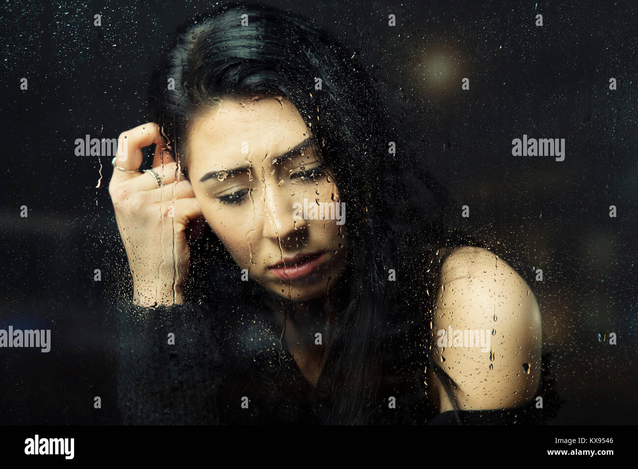 Woman sad depressed looking down hand on head sitting at rainy window ...