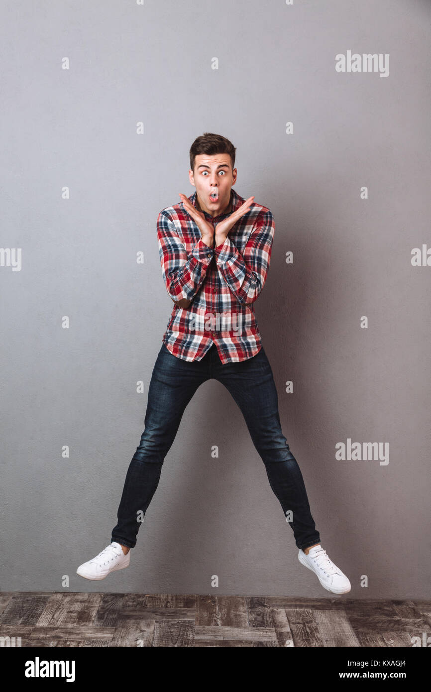 Full length image of Shocked man in shirt and jeans jumping and looking ...