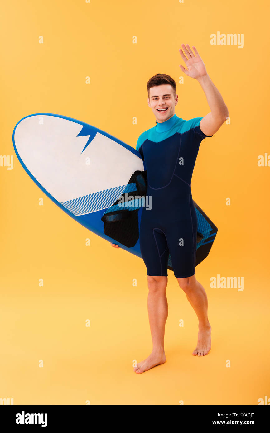 Full length image of happy surfer walking with surfboard and waving at ...