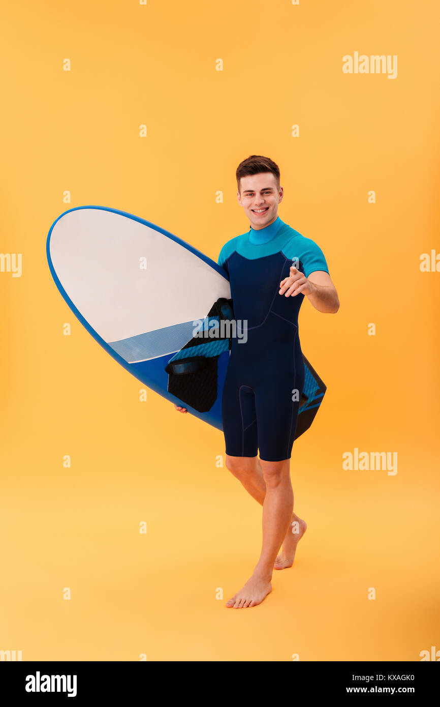 Full length image of smiling surfer walking with surfboard and pointing ...