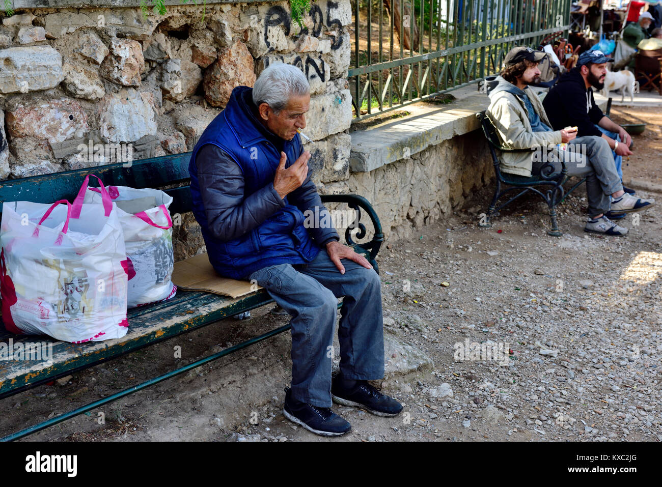Older Greek man relaxing, sitting outside on bench with shopping bags ...