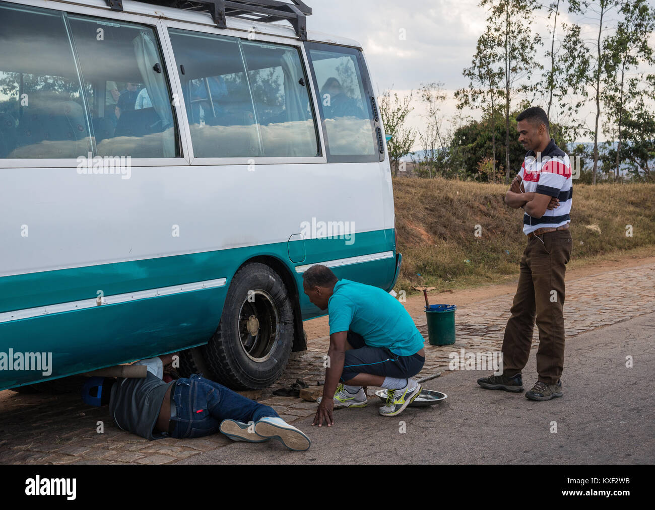 A man is under a minibus, trying to fix a flat tire, on the side of ...