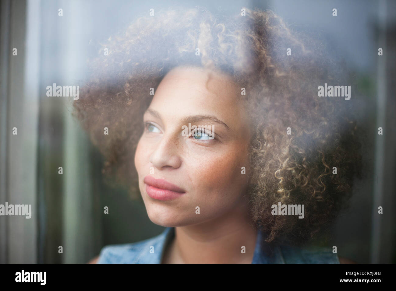 Close-up of thoughtful woman with curly hair seen through glass window ...