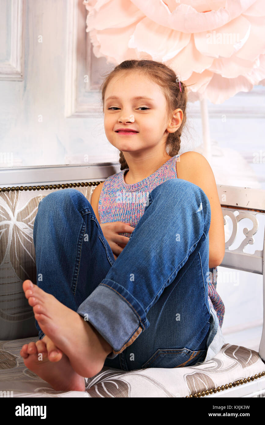 Beautiful laughing kid girl sitting on the bench in blue jeans and fashion blouse looking happy ...