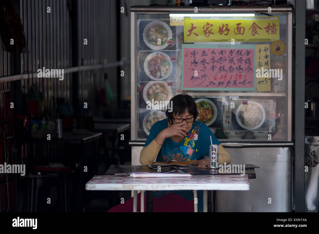 Elderly Asian woman sat at Chinese hawker restaurant table reading with ...