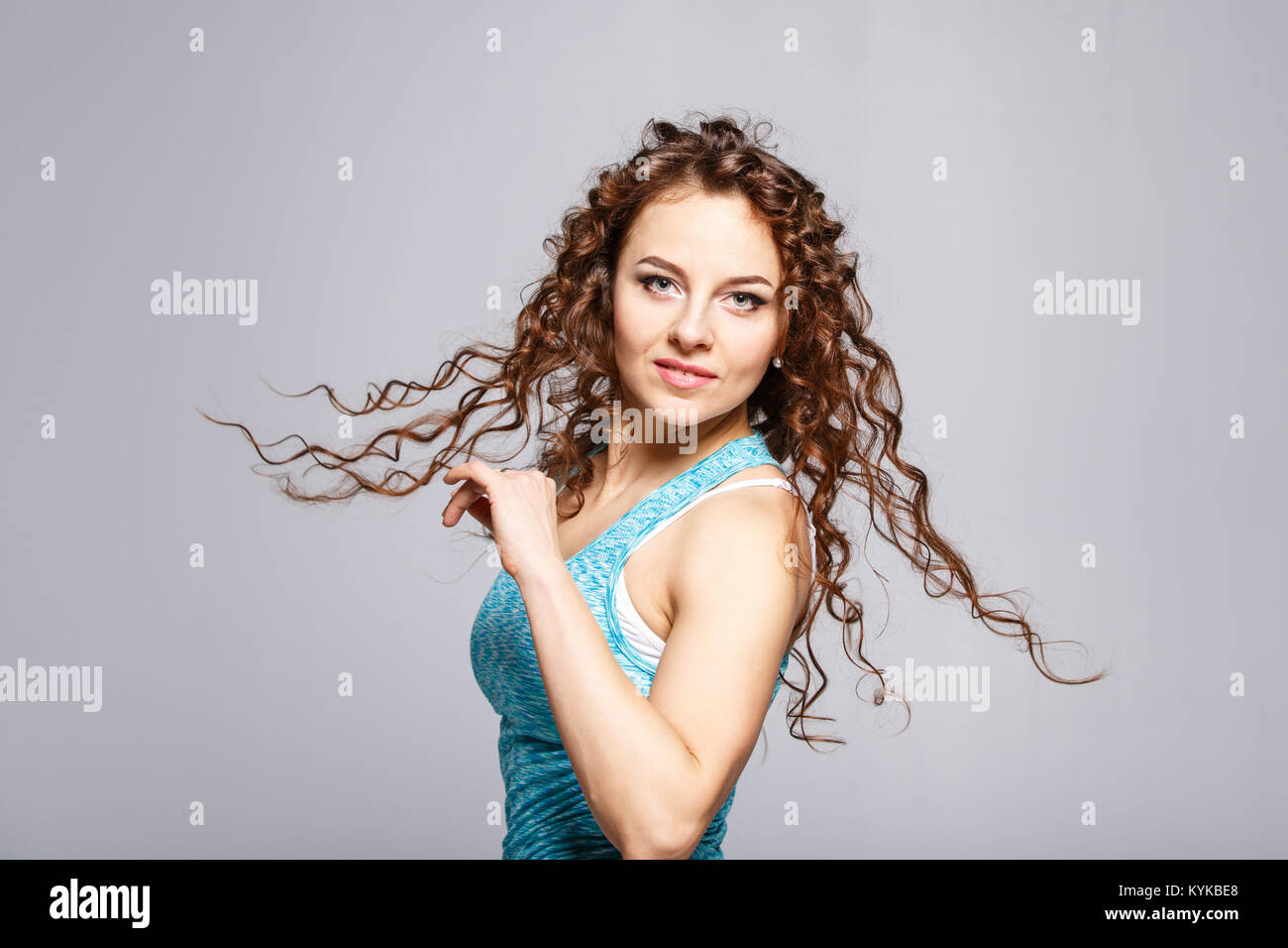 Young beautiful woman with curly windy hair dancing against grey wall ...