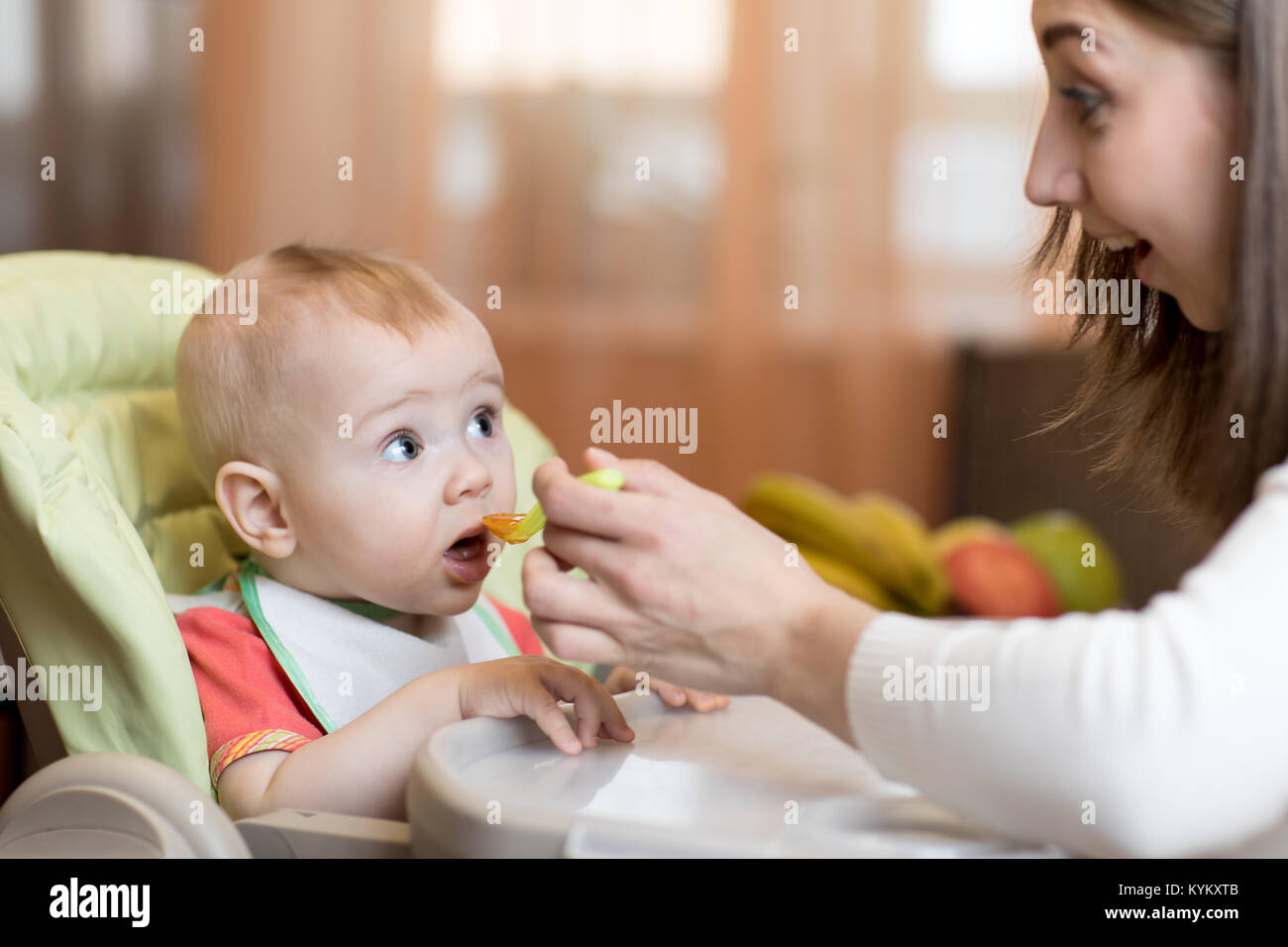 Mom giving homogenized food to her baby son on high chair in kitchen ...