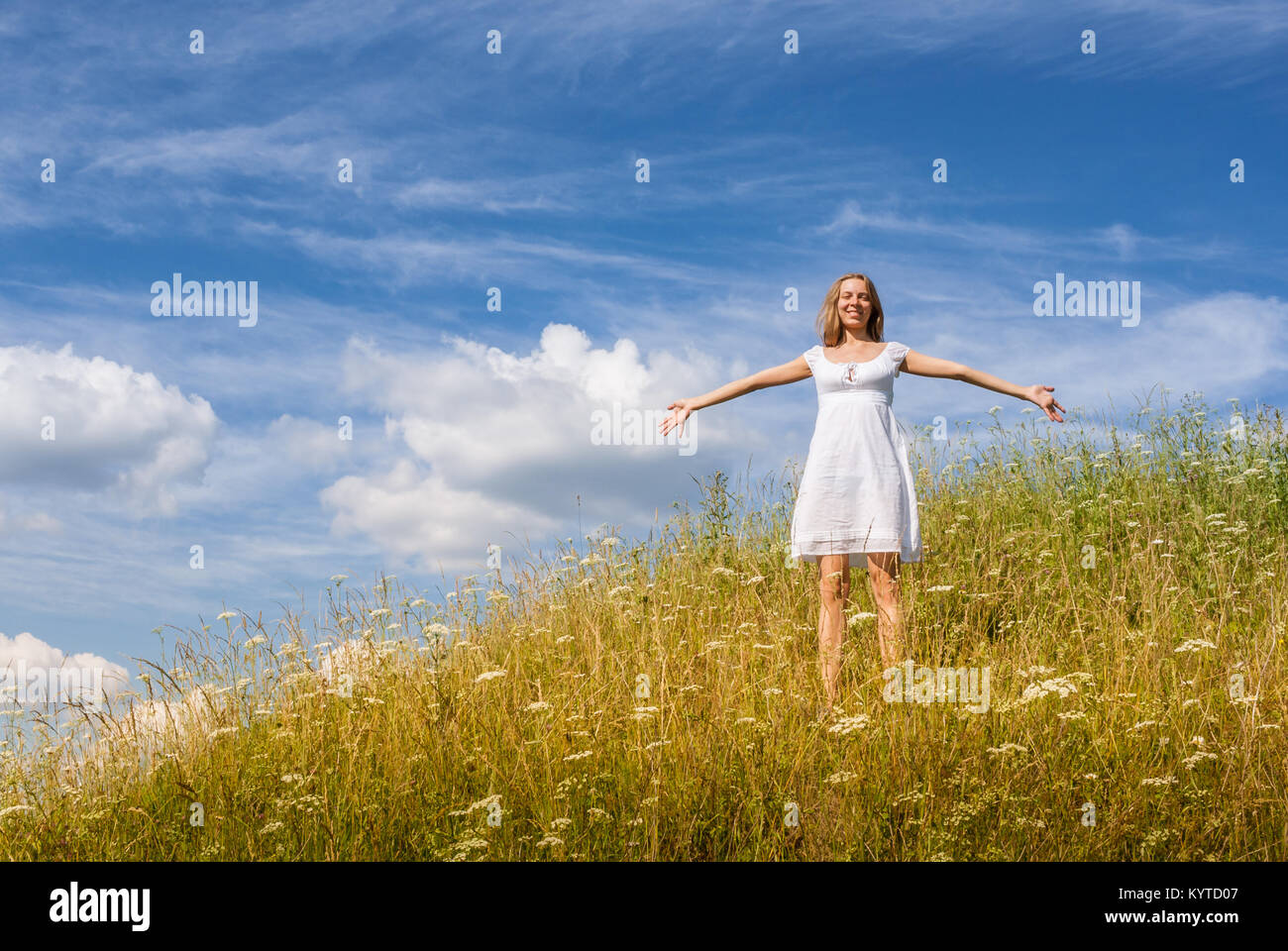 Young Girl In White Dress Is Standing On Grass Hill On Blue Sky