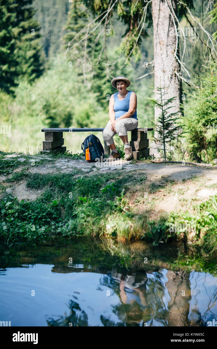 Woman sits on forest bench under tree, near water. Caucasian middle ...