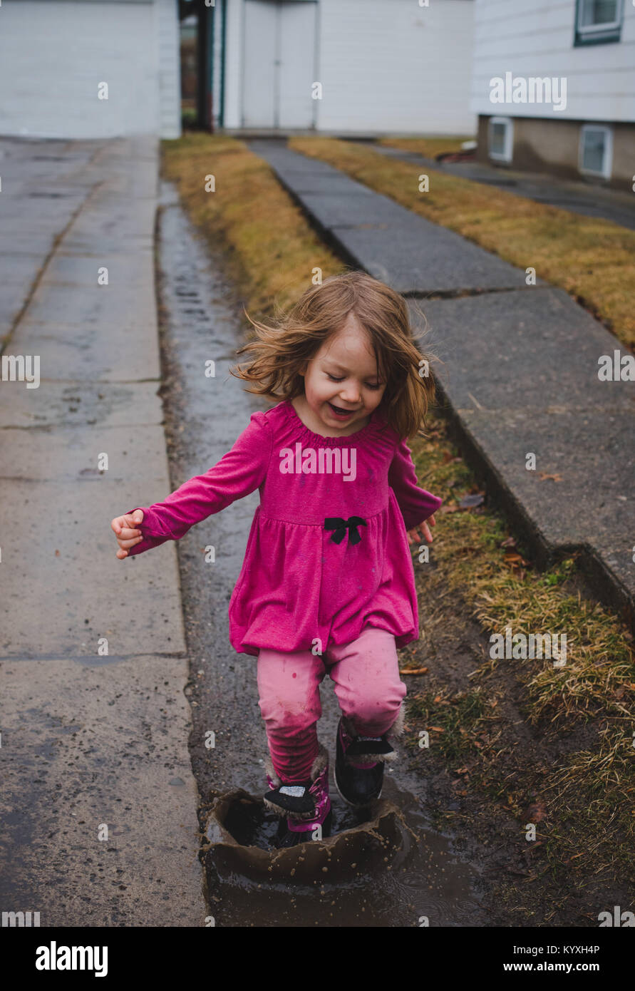 Toddler girl wearing pink, jumping in a mud puddle in a driveway on a ...