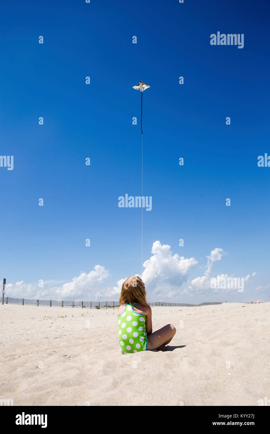 Rear view of girl flying kite while sitting at beach during sunny day ...