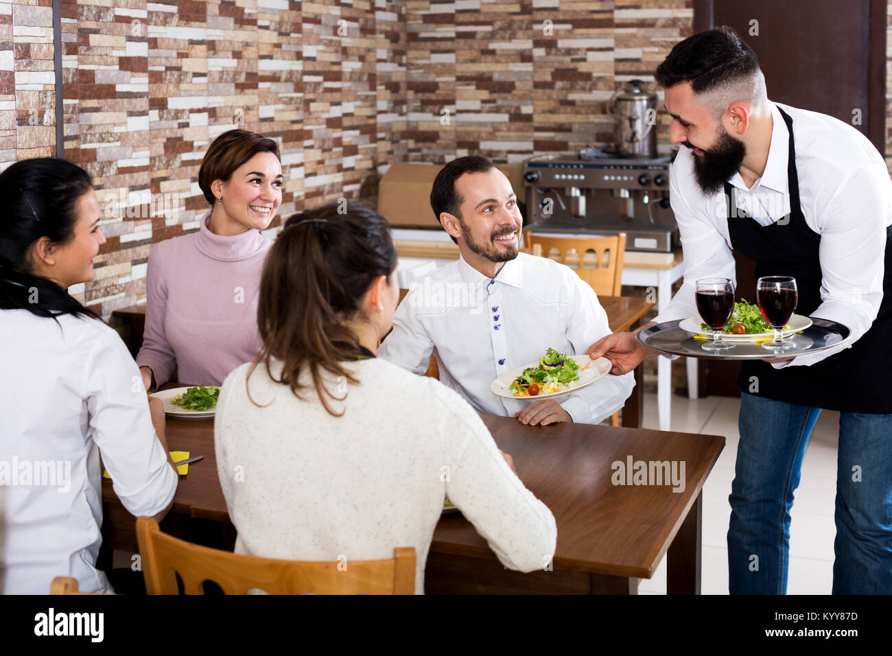 Smiling male waiter bringing order to visitors in country restaurant ...