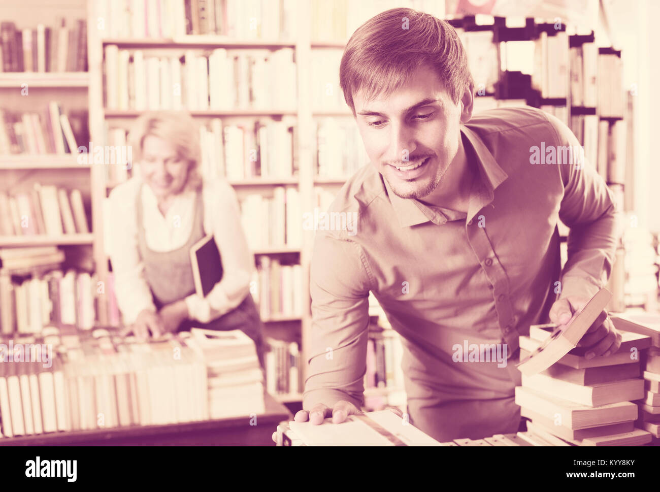 portrait of smiling young man standing among bookshelves and searching ...