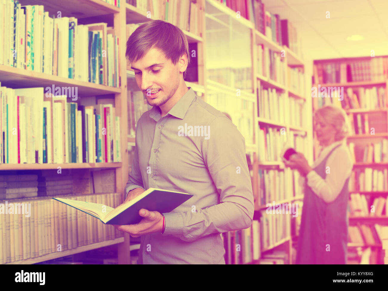 young man looking at open book standing among bookshelves Stock Photo ...
