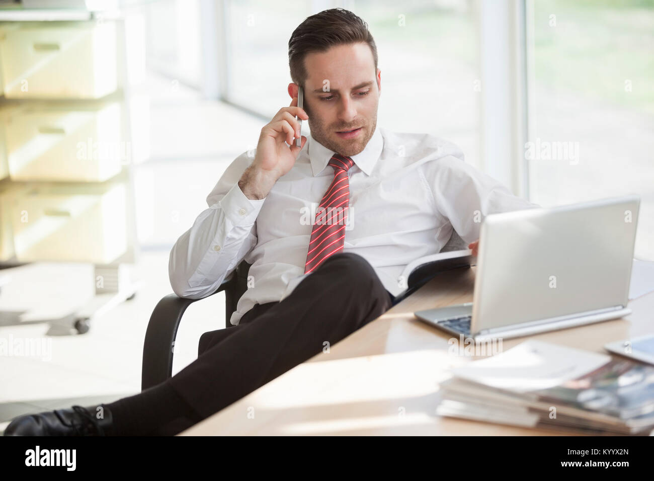 Young businessman using cell phone while reading book at office desk ...