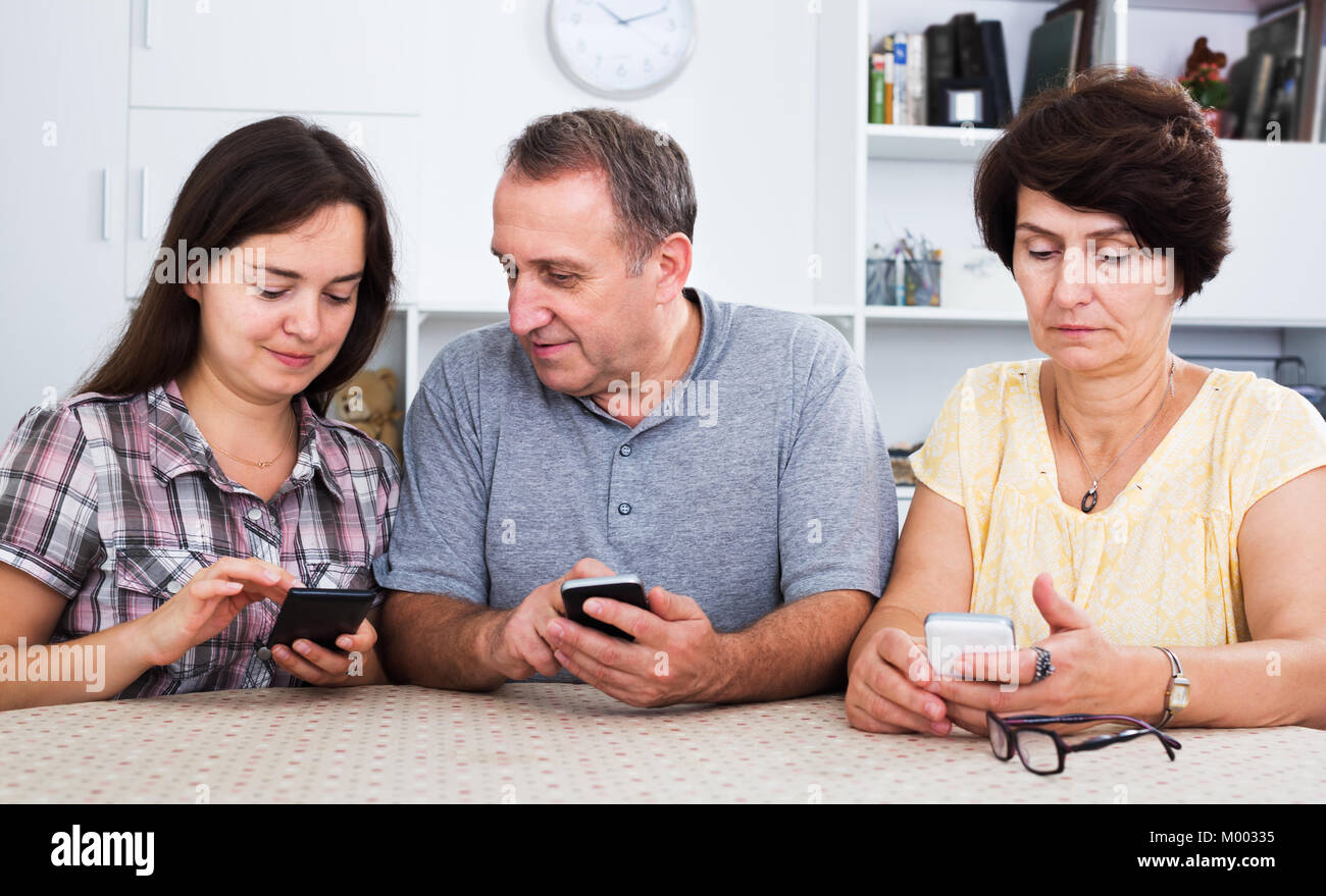 Cheerful elderly couple and young woman using mobile phones while sitting together at home ...