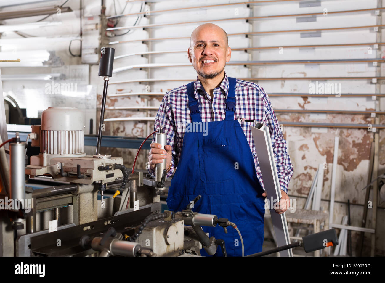 Smiling middle-aged worker in coverall toiling on a machine in PVC shop ...