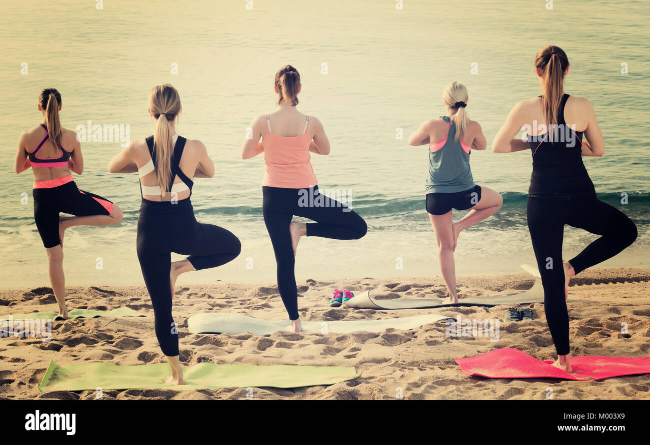 Back view of young females practicing various yoga positions on beach ...