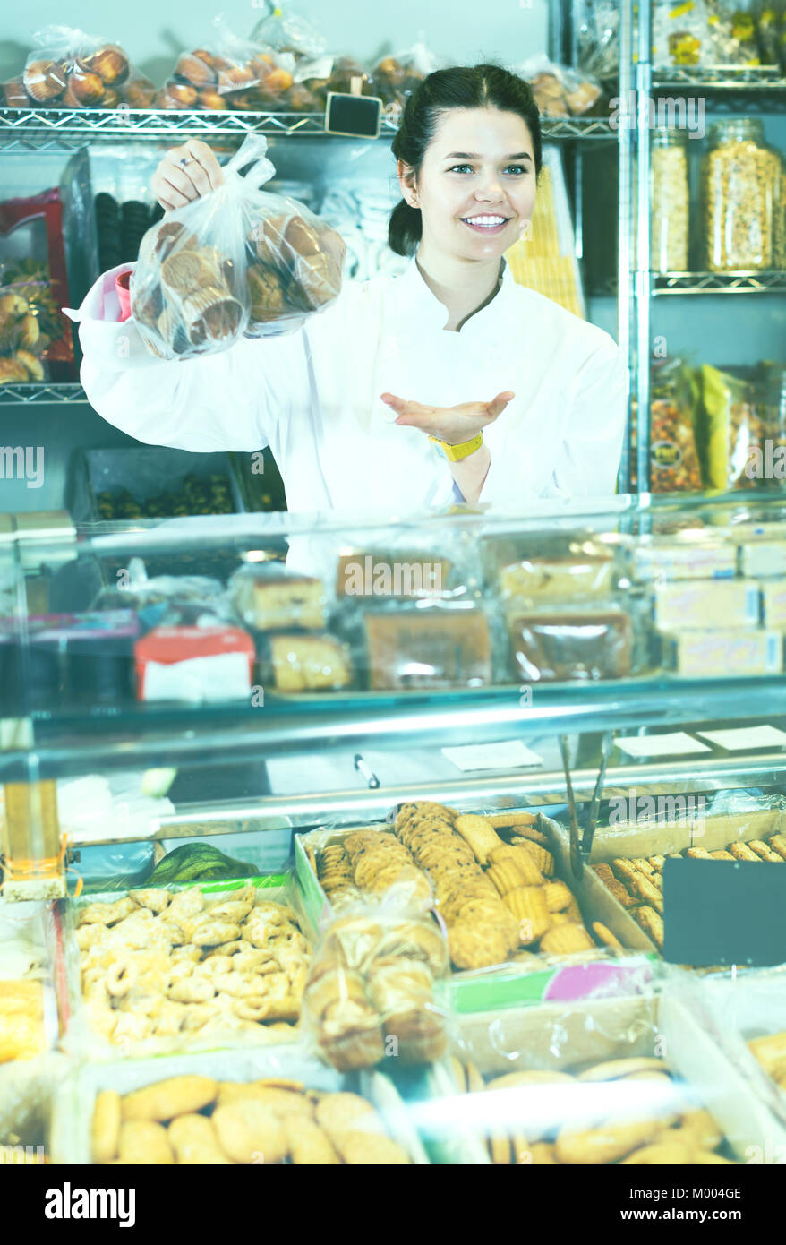 Young glad saleswoman posing with cookies at counter in candies shop ...
