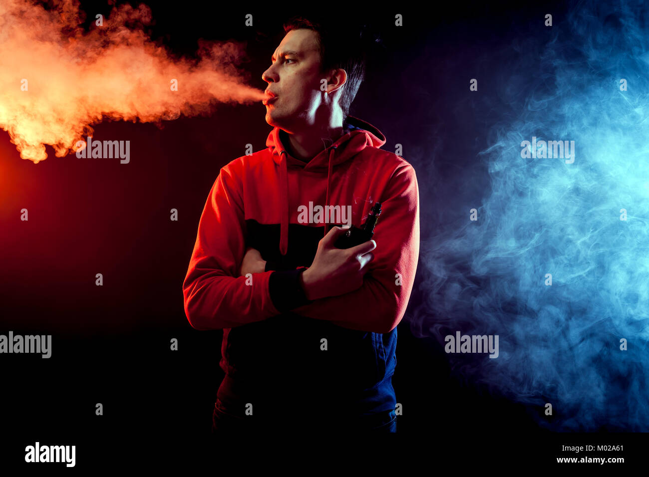 Young stylish male smoker in a sweatshirt is smoking a vape, and around ...