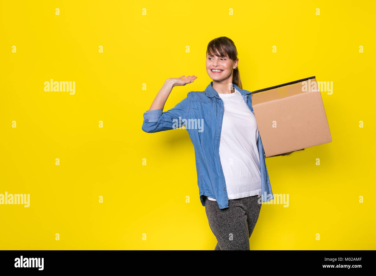 pretty woman standing on yellow background with moving cardboard box ...