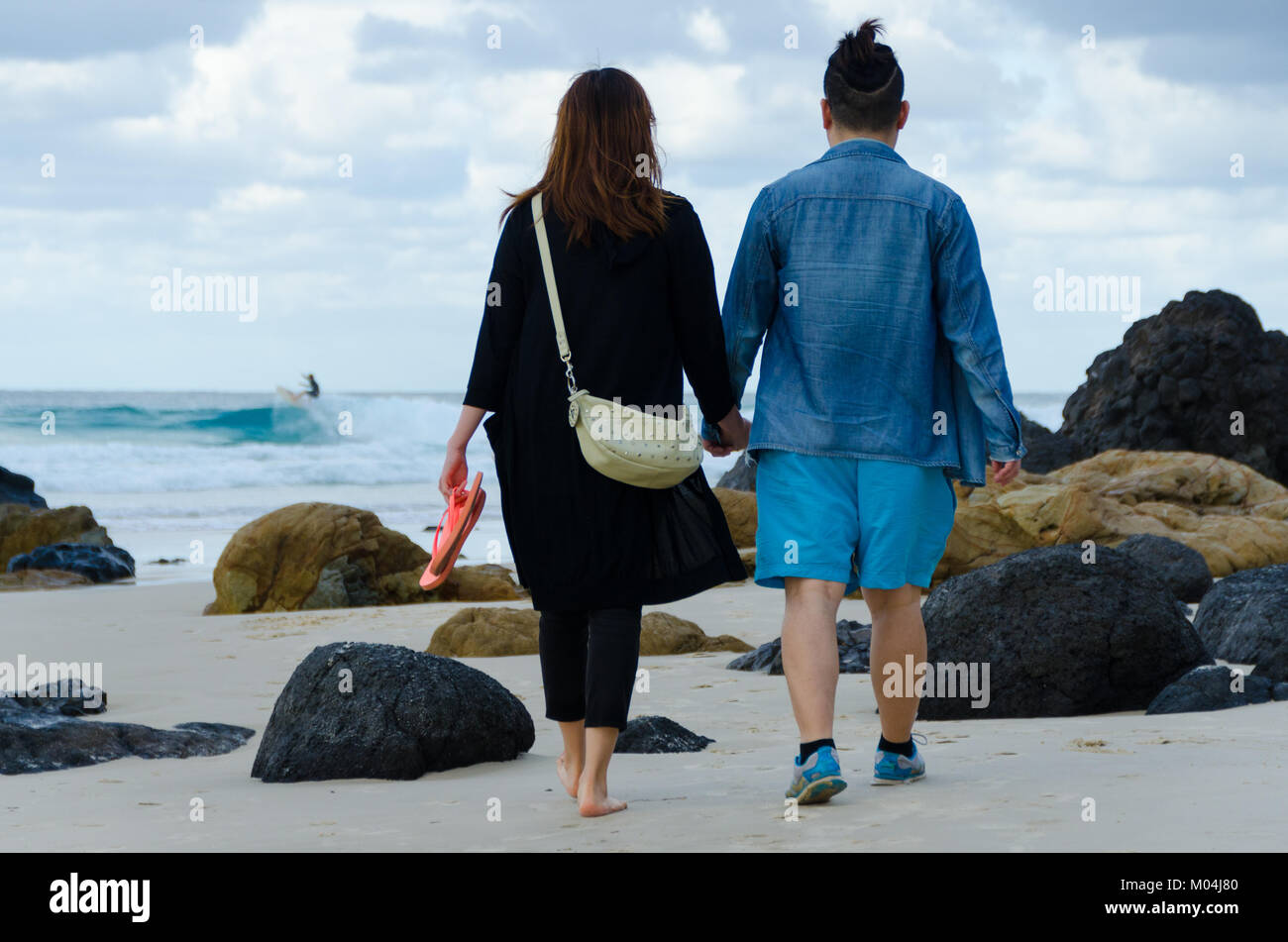 Couple walks across beach toward ocean with surfer riding wave in ...