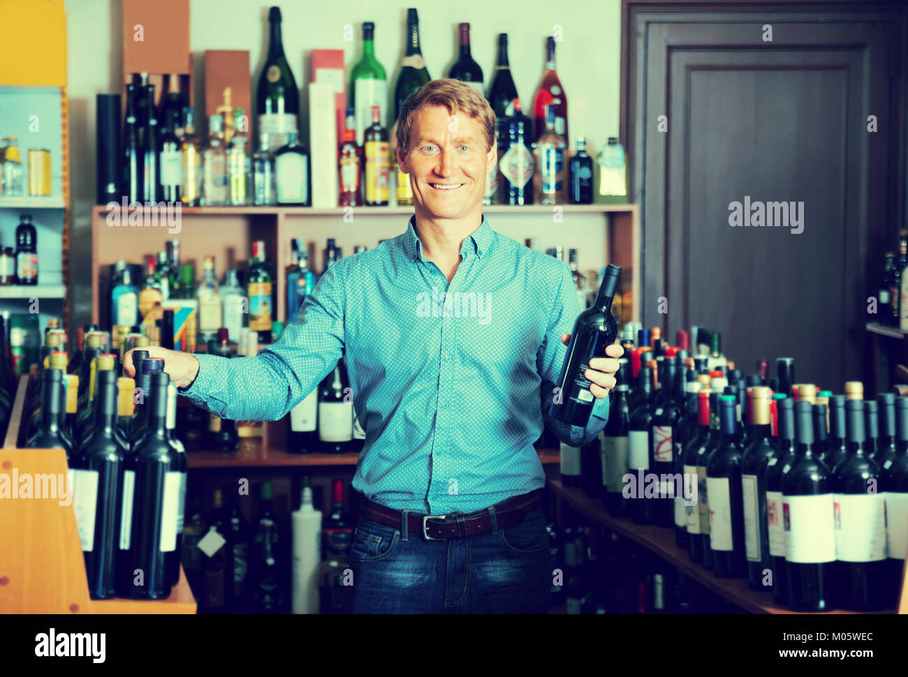 cheerful smiling man customer selecting bottle of wine in wine store ...