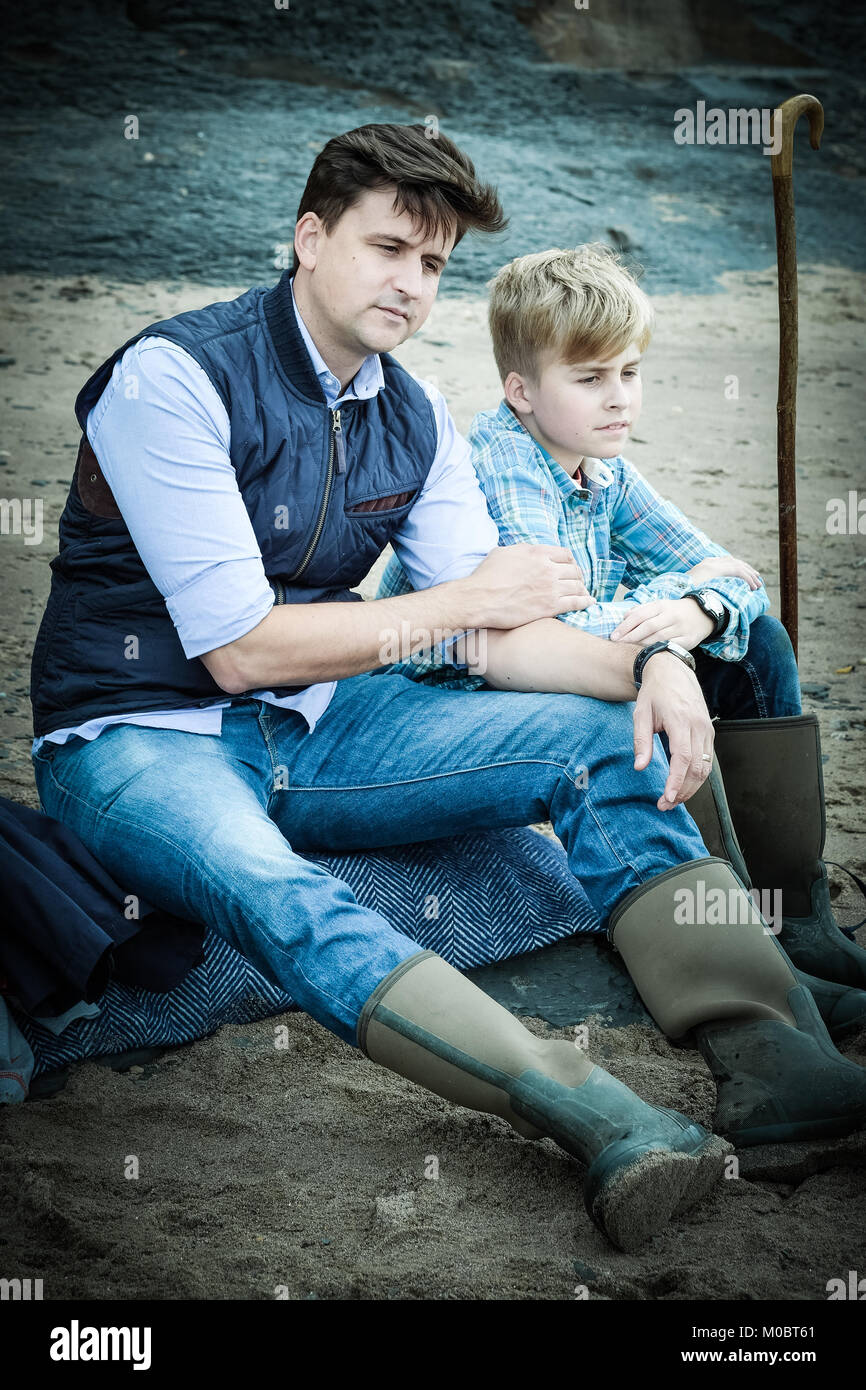 Father and son sitting on rocks at the beach, looking thoughtful, sad ...