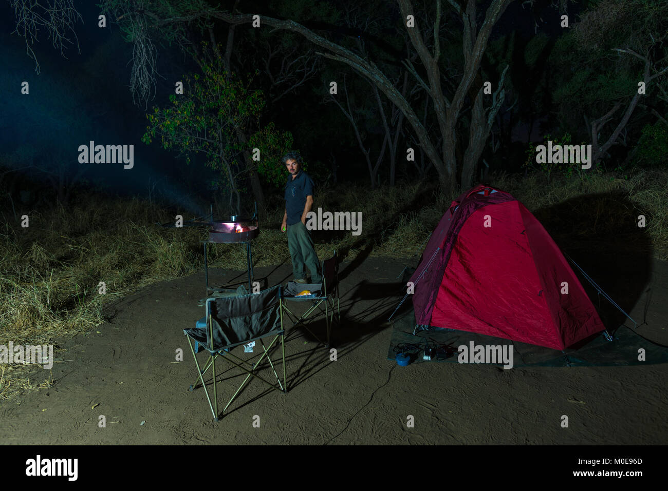 Man cooking with fire wood and braai equipment by night. Tent and ...