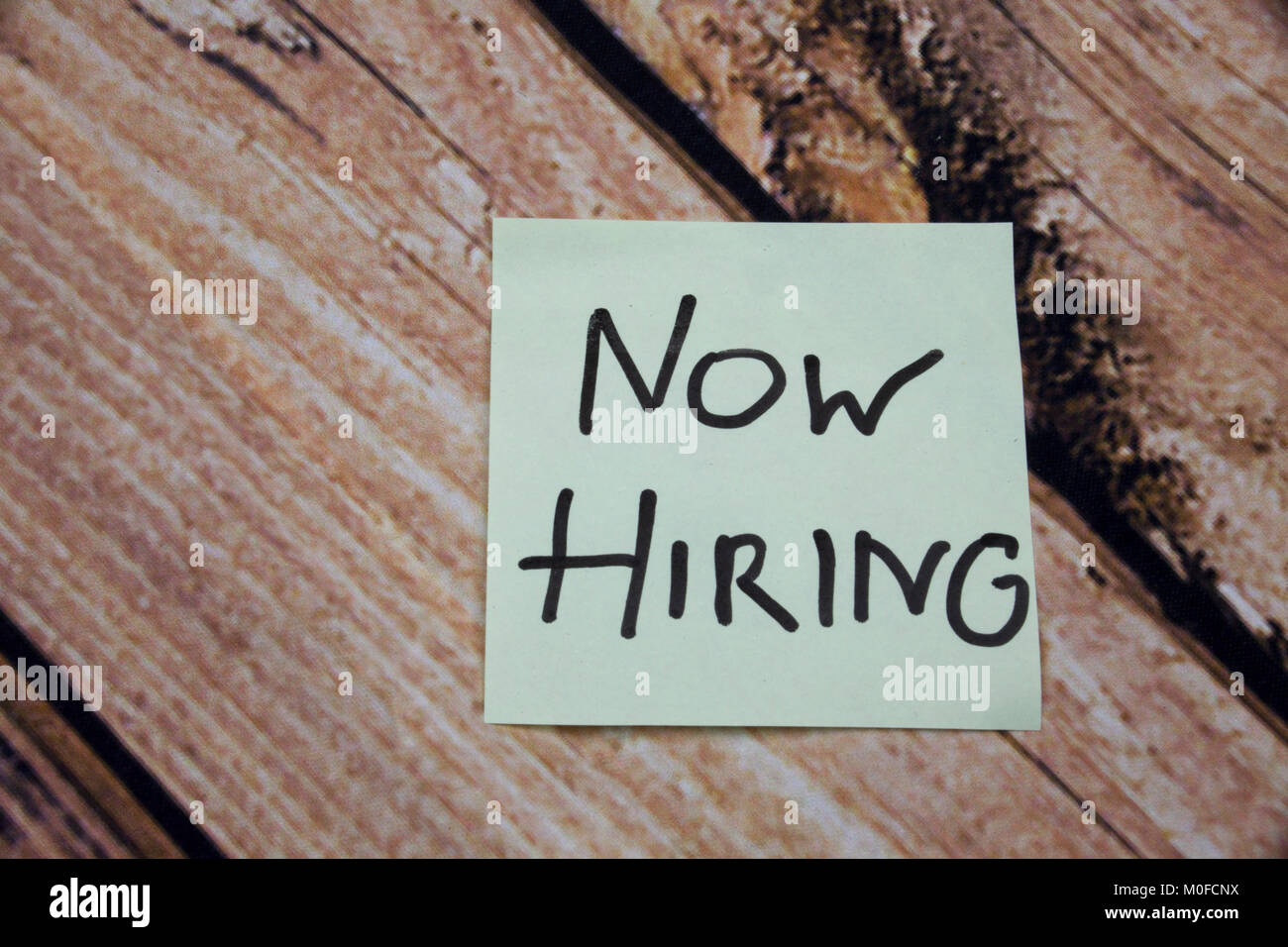 Now Hiring written on an yellow sticky note paper on the wooden oak ...