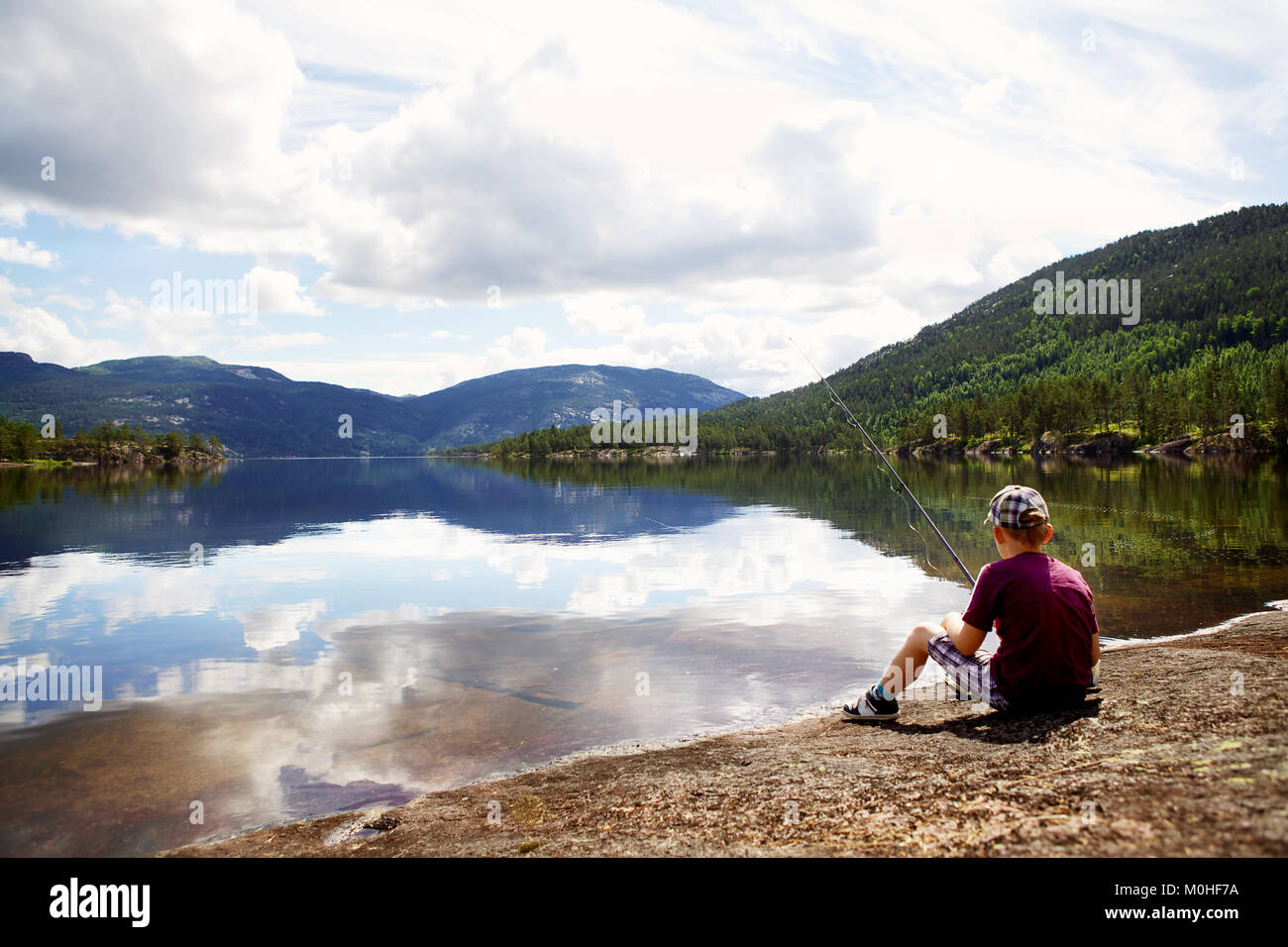 A boy is sitting and fishing in a lake with clouds reflection on water ...