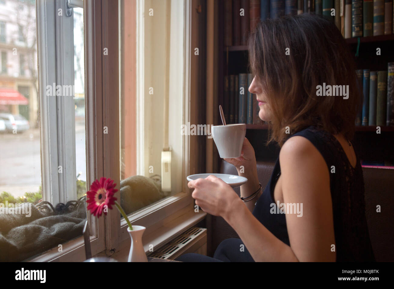 young woman with cup of coffee sitting by the window with books in the ...