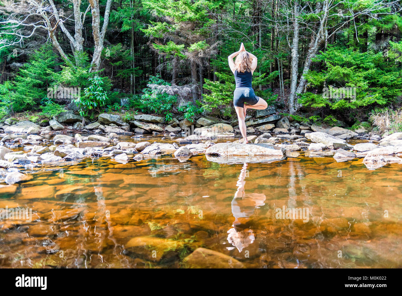 Young woman in tree yoga pose standing on one leg on rock by river in ...