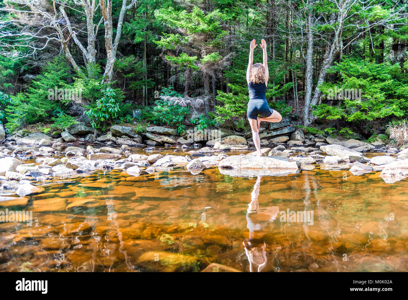 Young woman in tree yoga pose standing on one leg on rock by river in ...