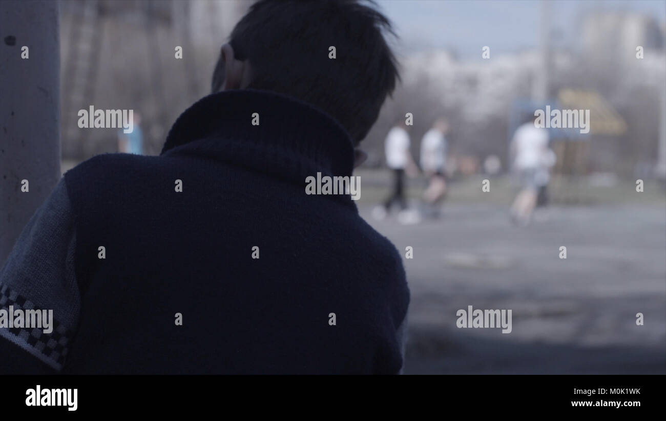 Lonely boy sits on the bench and watches as his friends play football ...