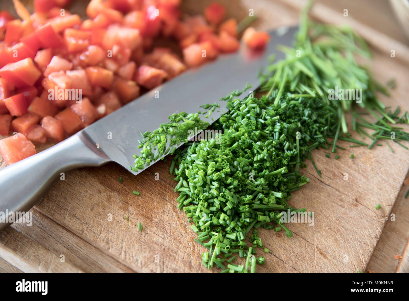 Chopped chives on cutting board with chef's knife and chopped tomatoes ...