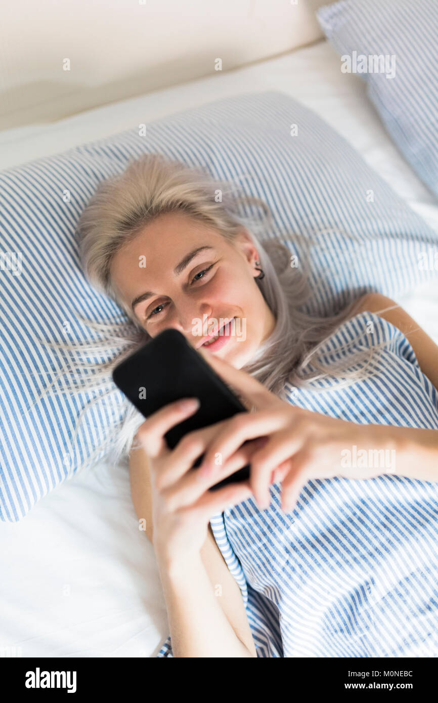 Smiling young woman lying in bed checking cell phone Stock Photo - Alamy