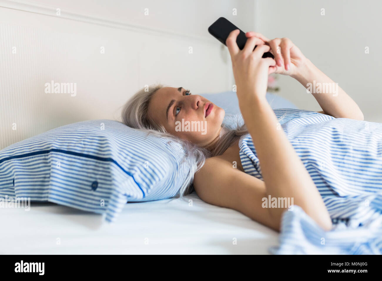 Serious young woman lying in bed checking cell phone Stock Photo - Alamy