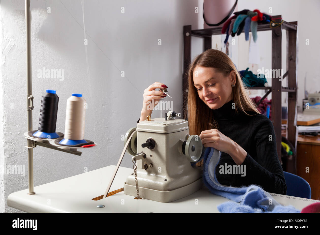 Young woman Furrier sewing blue fur coat on sewing machine in workshop ...