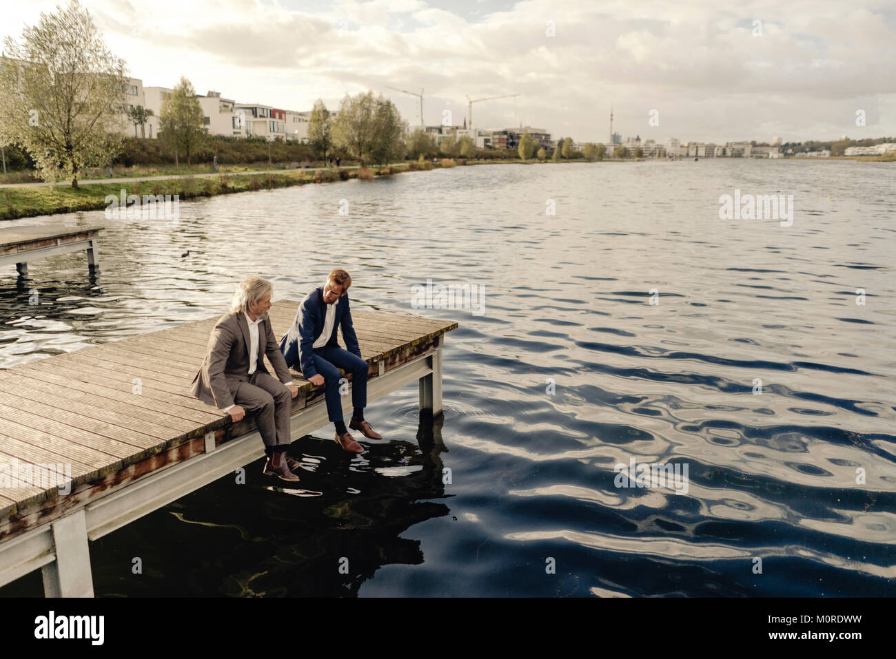 Two businessmen sitting on jetty at a lake talking Stock Photo - Alamy