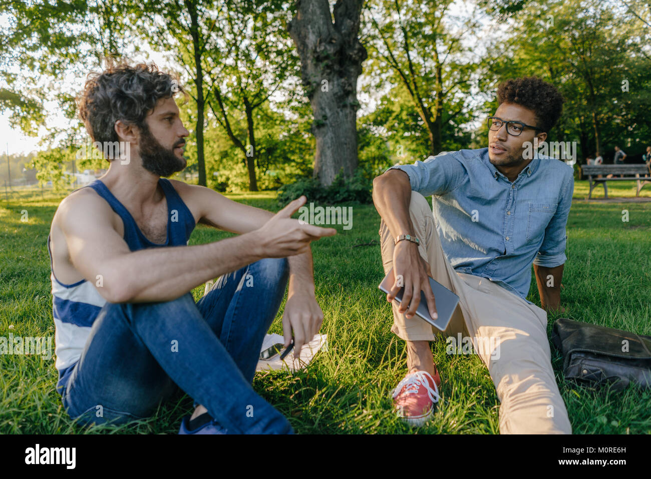 Two friends sitting in a park with mobile device and papers discussing ...